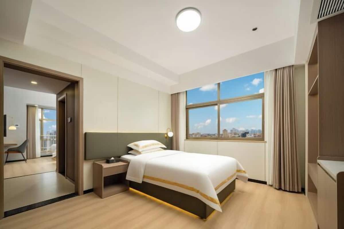 Quarto de hotel com cama, mesa de cabeceira, vista da cidade e porta para uma área de estar.