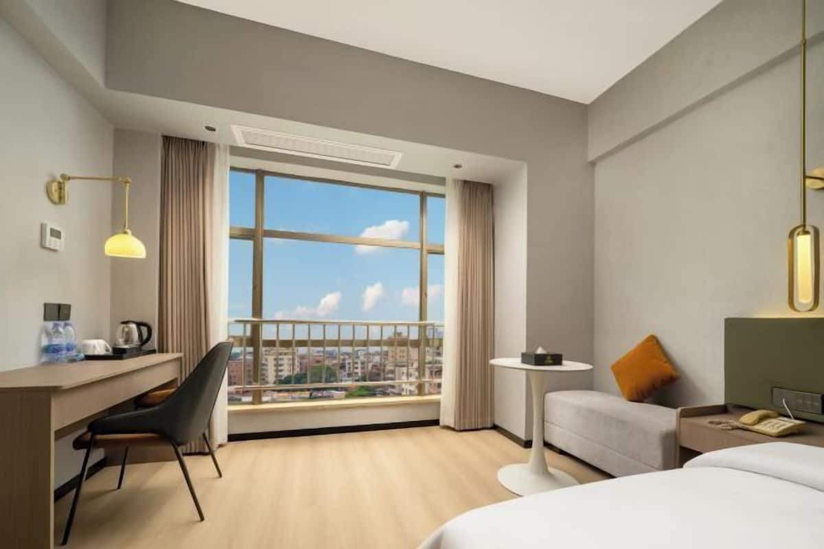 Quarto de hotel com escrivaninha, cadeira, cama, mesa de cabeceira, abajur e vista da cidade através de amplas janelas.