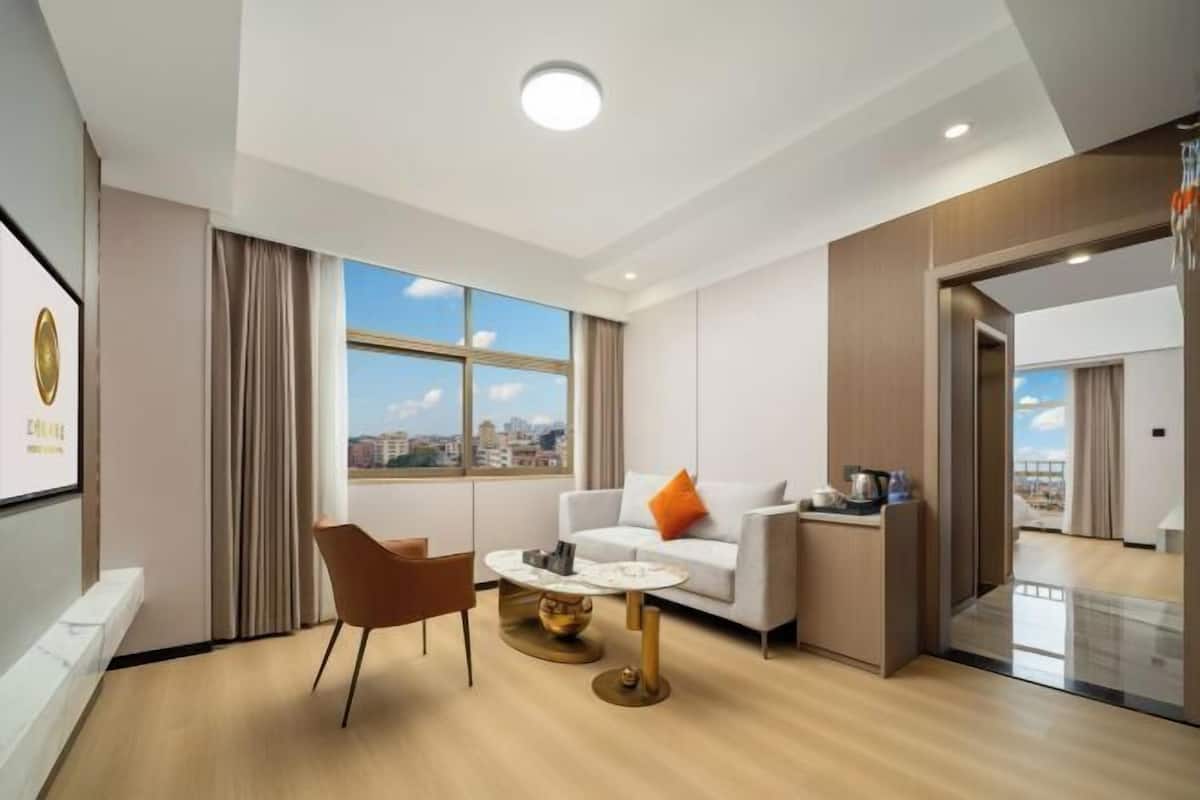 Quarto de hotel moderno com sofá, poltrona e mesa de centro, grandes janelas com vista para a cidade e piso de madeira.