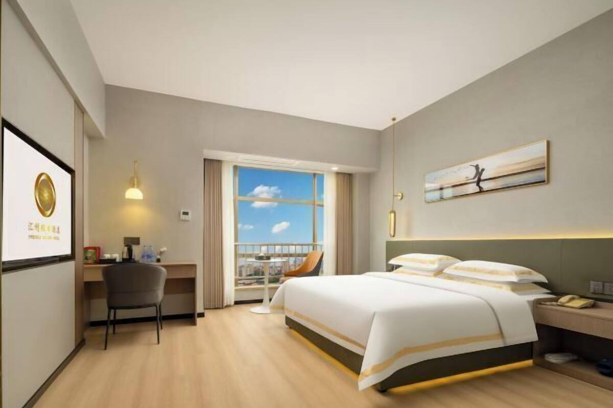 Quarto de hotel com cama, escrivaninha, cadeira e vista da cidade através de uma grande janela.