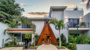 Um edifício moderno com fachada triangular de madeira, uma varanda com assentos e vegetação exuberante ao redor.