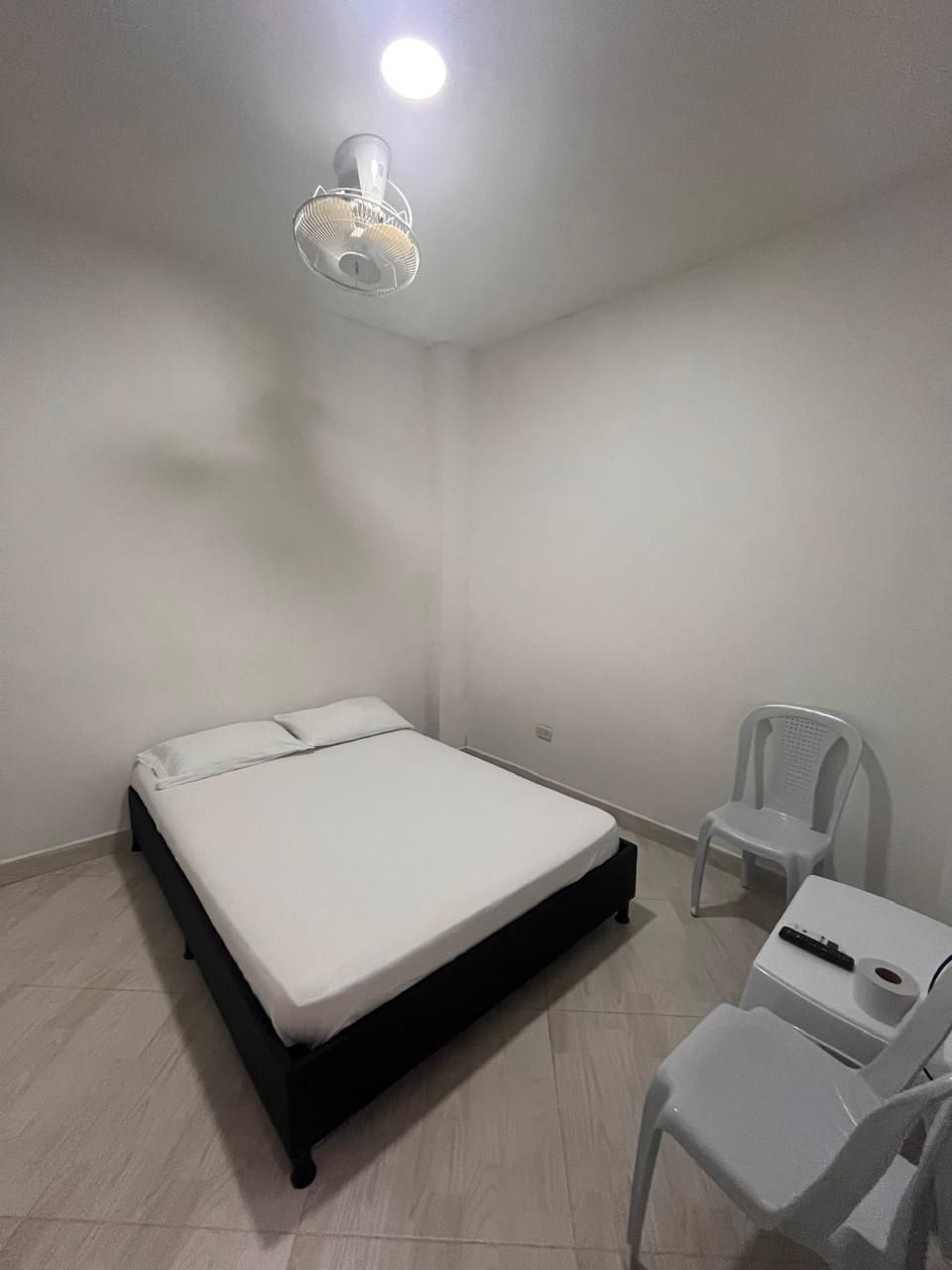 Quarto pequeno e limpo, com uma cama de solteiro, uma escrivaninha, duas cadeiras e um ventilador de teto.