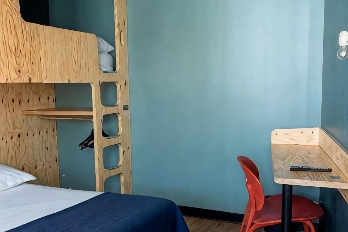 Uma cama beliche com escada de madeira, uma cama de solteiro com colcha azul, uma pequena escrivaninha de madeira com cadeira e uma parede azul claro.