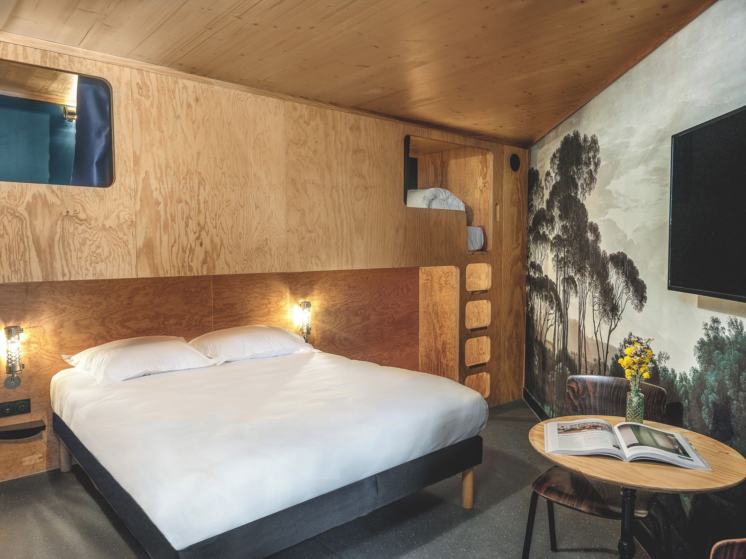 Um quarto de madeira com uma cama, uma pequena mesa com um livro e uma televisão fixada na parede.
