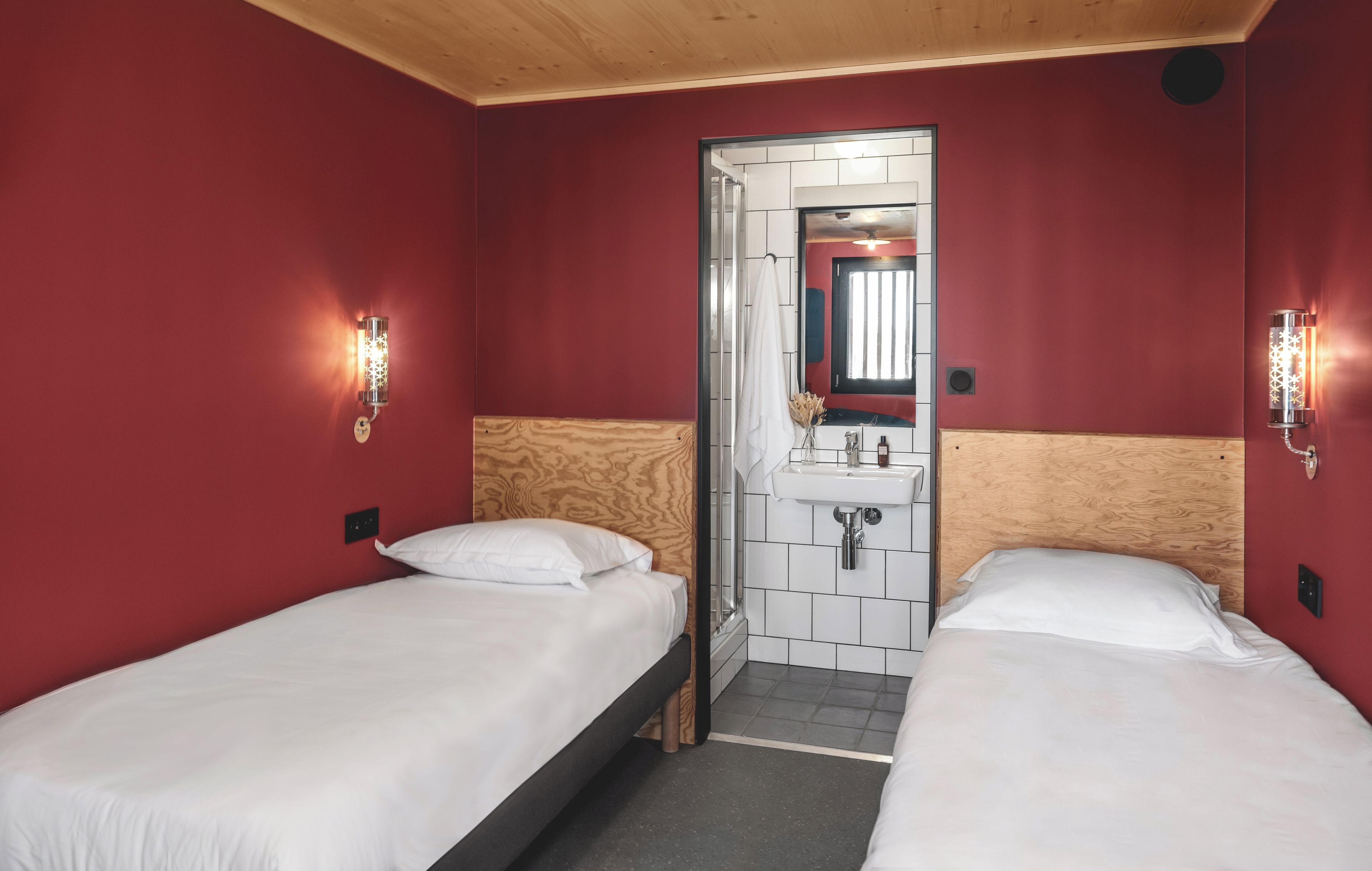 Quarto pequeno e compacto com duas camas de solteiro, cabeceira de madeira, pia pequena e espelho acima.