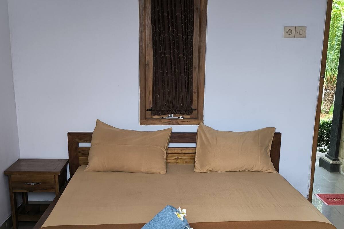 Quarto com cama, duas almofadas, cabeceira de madeira, mesa de cabeceira, janela com cortinas e porta que dá para o exterior.