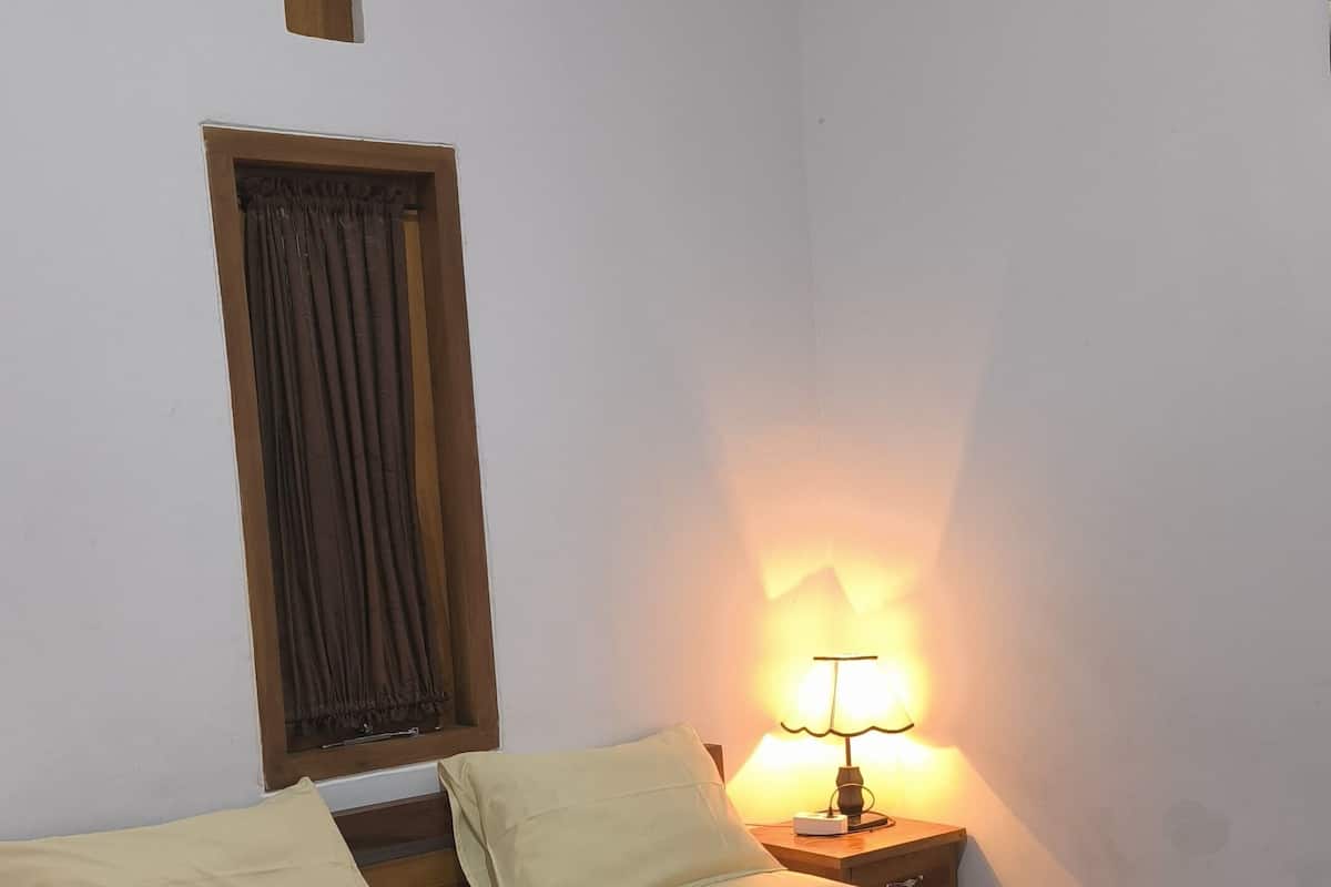 Quarto com cama, mesa de cabeceira com abajur, janela com cortinas e uma pequena estante de madeira.