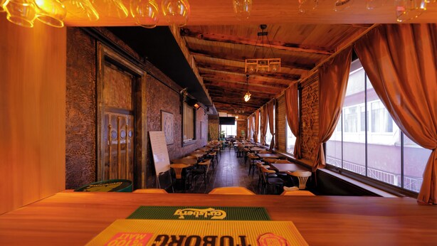 Restaurante com mesas compridas, luminárias pendentes e grandes janelas com cortinas.