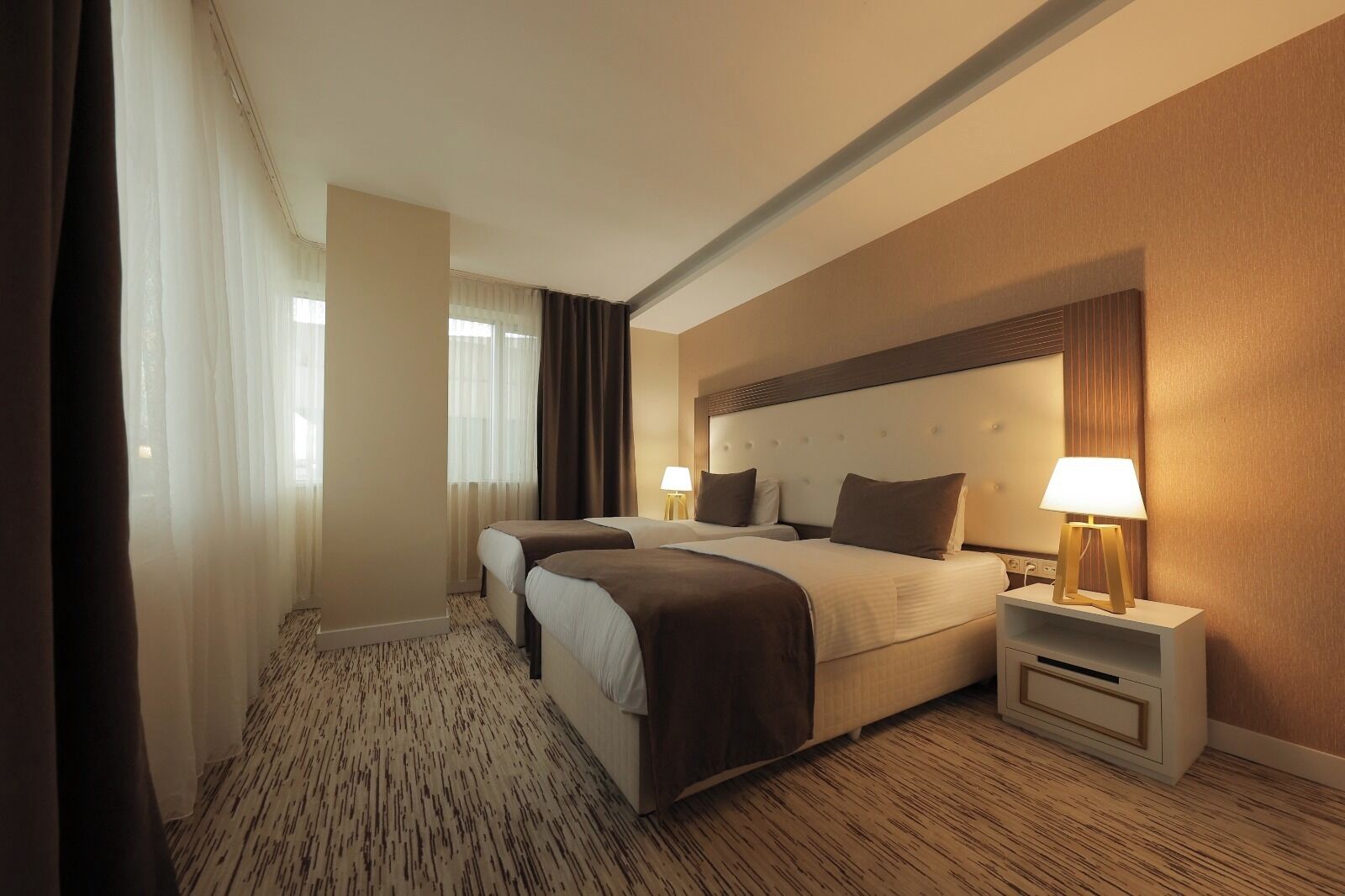 Quarto de hotel com duas camas, uma mesa de cabeceira com abajur, uma janela grande com cortinas e um tapete estampado.