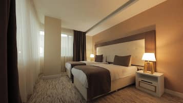 Quarto de hotel com duas camas, uma mesa de cabeceira com abajur, uma janela grande com cortinas e um tapete estampado.