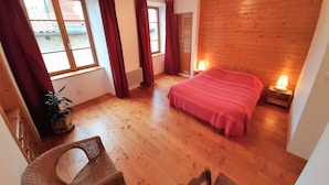Quarto com piso de madeira, cama com cobertor vermelho, duas luminárias de cabeceira, janela com cortinas e uma cadeira.