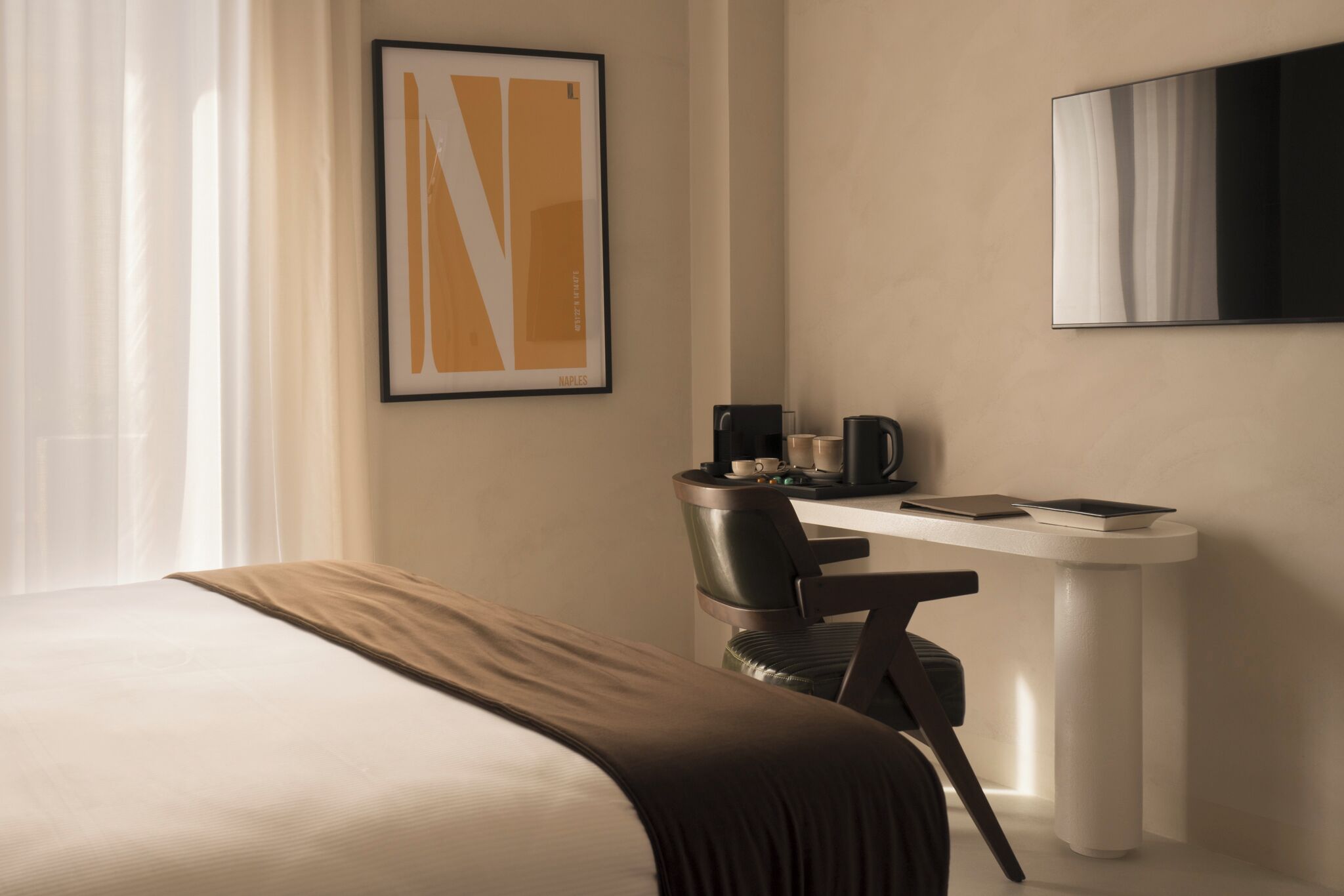 Quarto de hotel com cama, escrivaninha com cadeira, máquina de café e uma obra de arte emoldurada na parede.