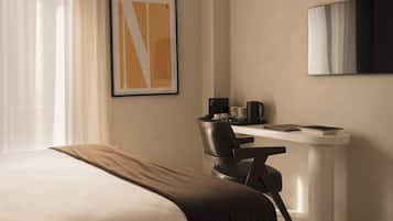 Quarto de hotel com cama, escrivaninha com cadeira, máquina de café e uma obra de arte emoldurada na parede.