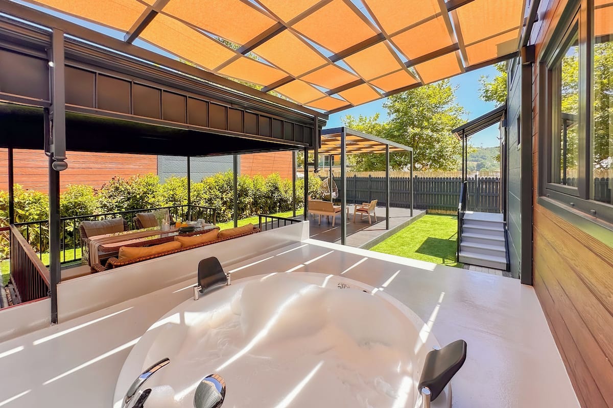 Uma área moderna para hidromassagem ao ar livre, com pérgola, deck de madeira e vista para um quintal com gramado e cerca de madeira.