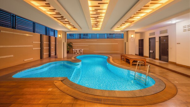An indoor swimming pool with a wooden deck surrounding it, a seating area with a table and chairs, and a ceiling with recessed lighting.