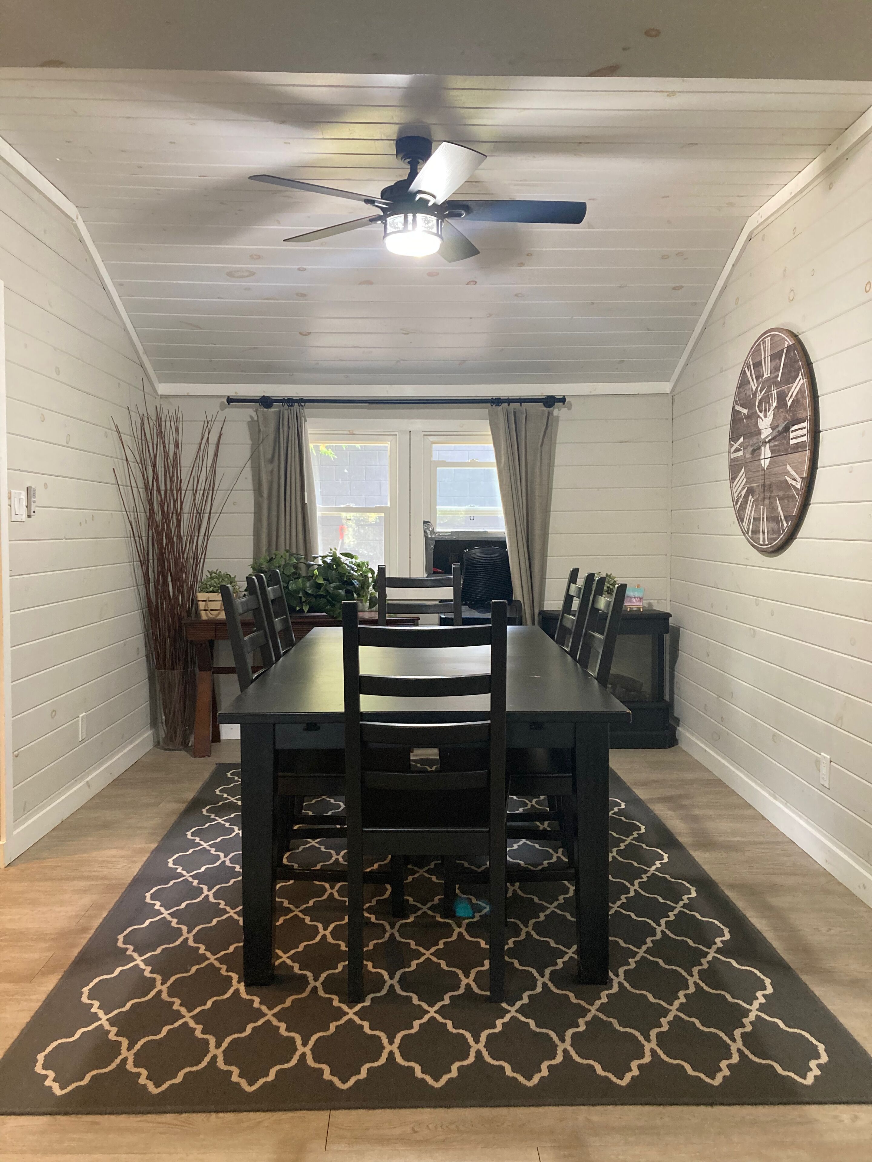 Uma sala de jantar com mesa de madeira, cadeiras, ventilador de teto e um relógio de parede grande.