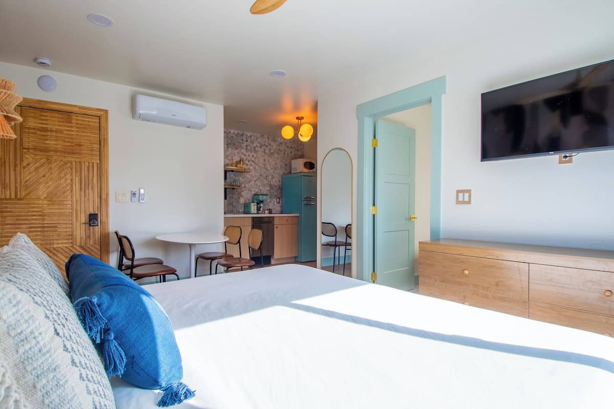 Quarto com cama, cômoda de madeira, televisão e cozinha compacta ao fundo.