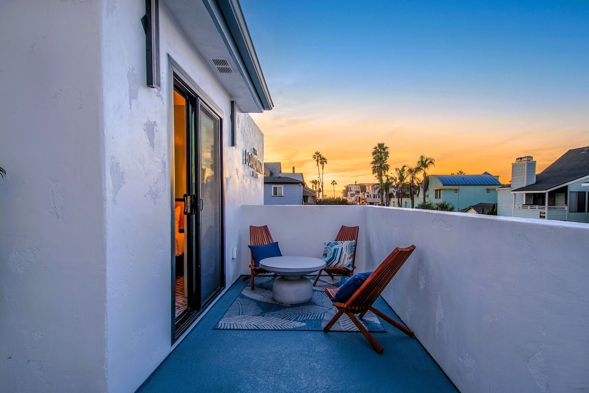 Varanda com mesa redonda e duas cadeiras, porta de vidro deslizante e vista para palmeiras e outros edifícios ao pôr do sol.