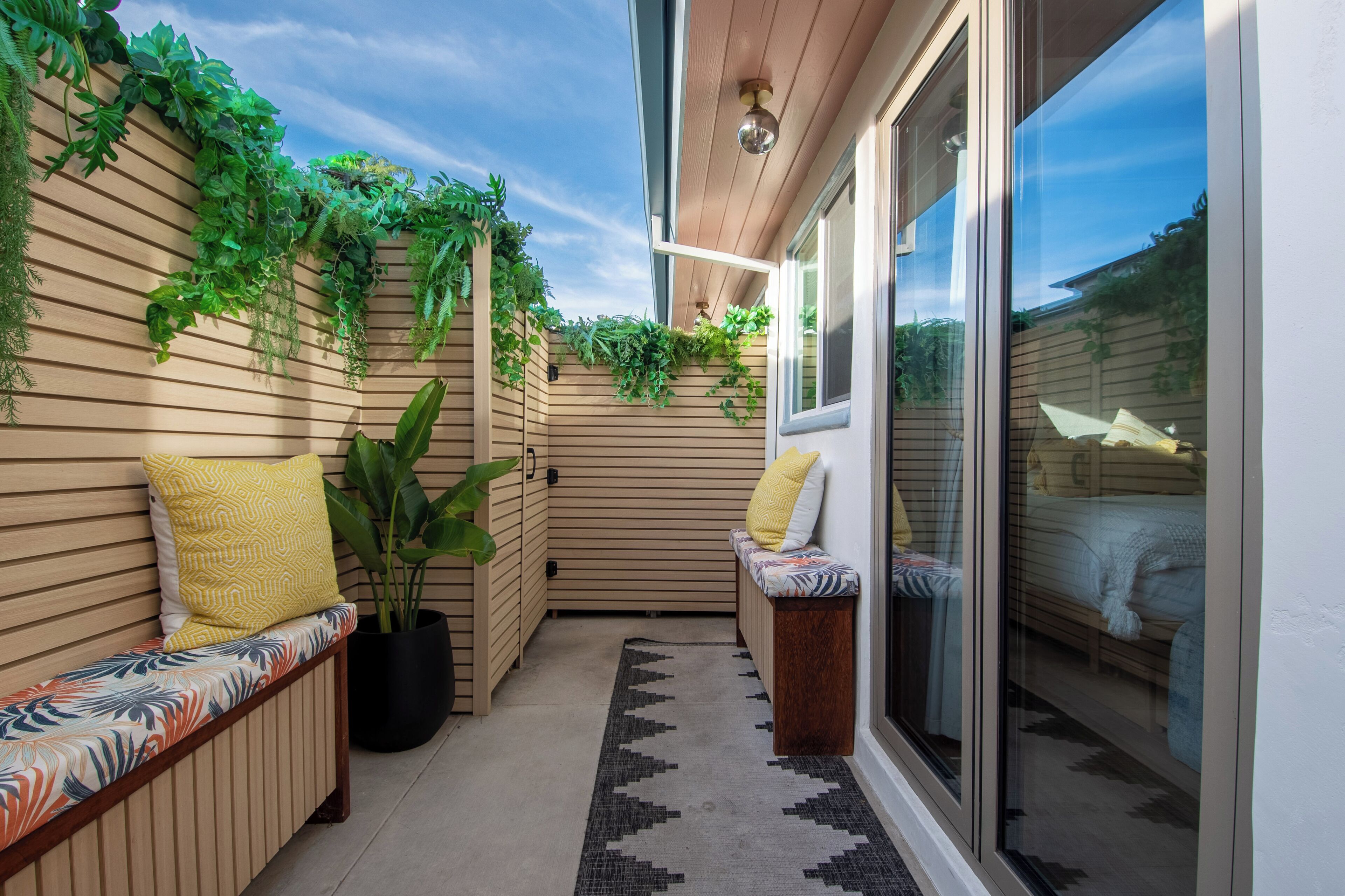 Uma área externa moderna com bancos de madeira, uma planta em vaso e uma porta de vidro que dá para um quarto.