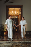 Um casal de mãos dadas em um hall iluminado, com um lustre e um vaso de planta.