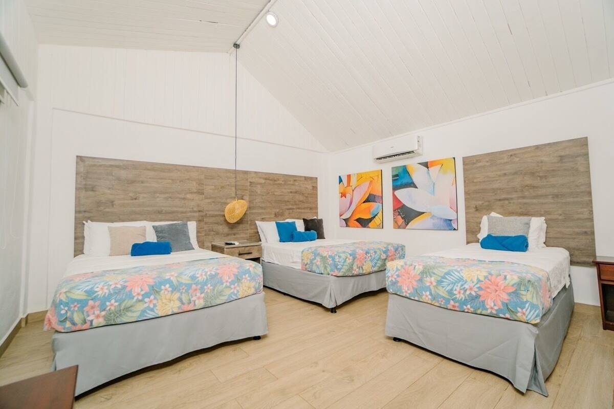 Quarto com três camas, cada uma com roupa de cama florida, cabeceiras de madeira e arte abstrata nas paredes.