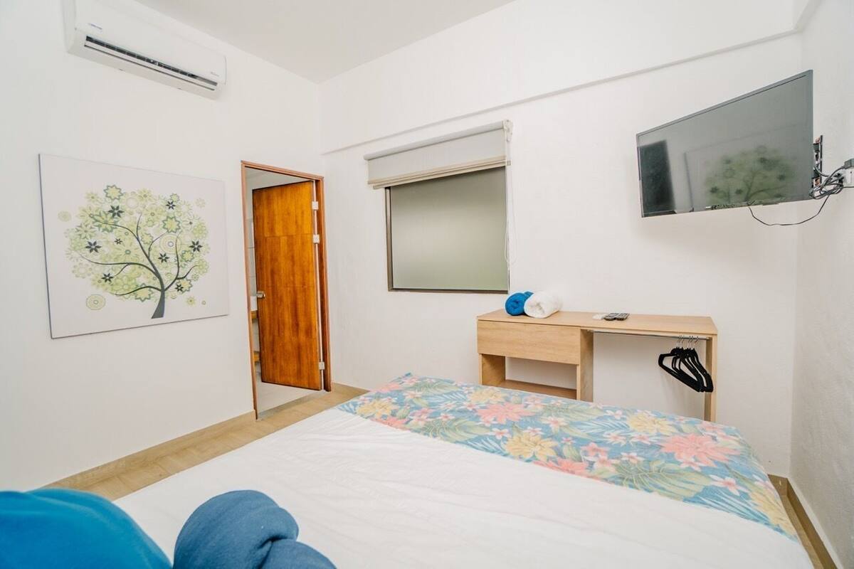Quarto com cama, escrivaninha, espelho e televisão fixada na parede.