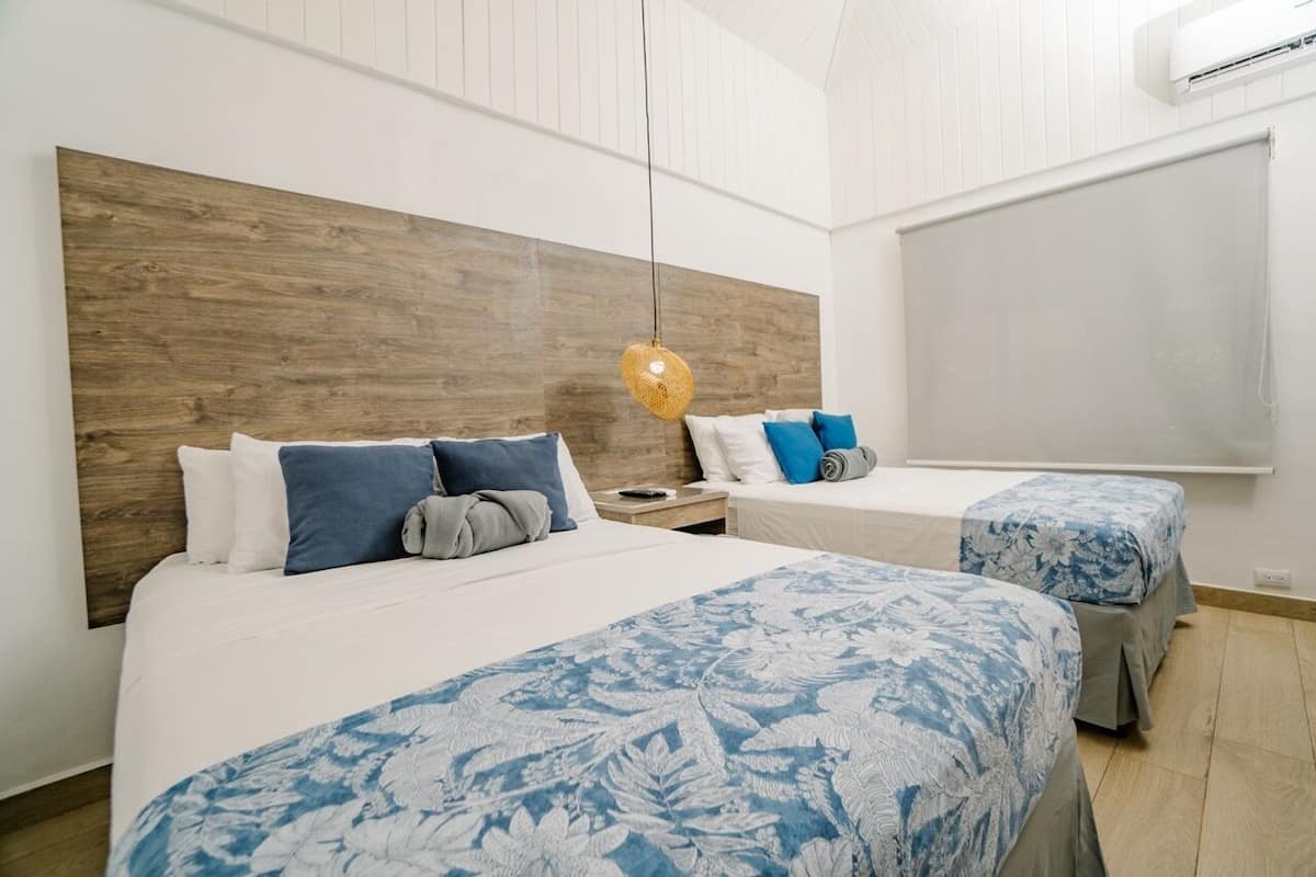 Quarto de hotel com duas camas, uma cabeceira de madeira, luminária pendente e uma janela grande.