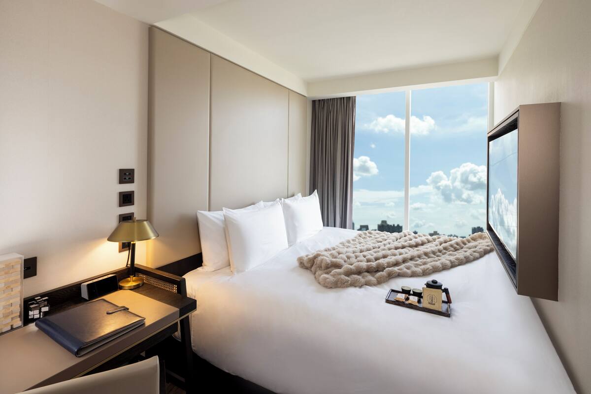 Quarto de hotel com uma cama grande, uma escrivaninha com candeeiro, vista para a cidade pela janela e uma TV fixada na parede.