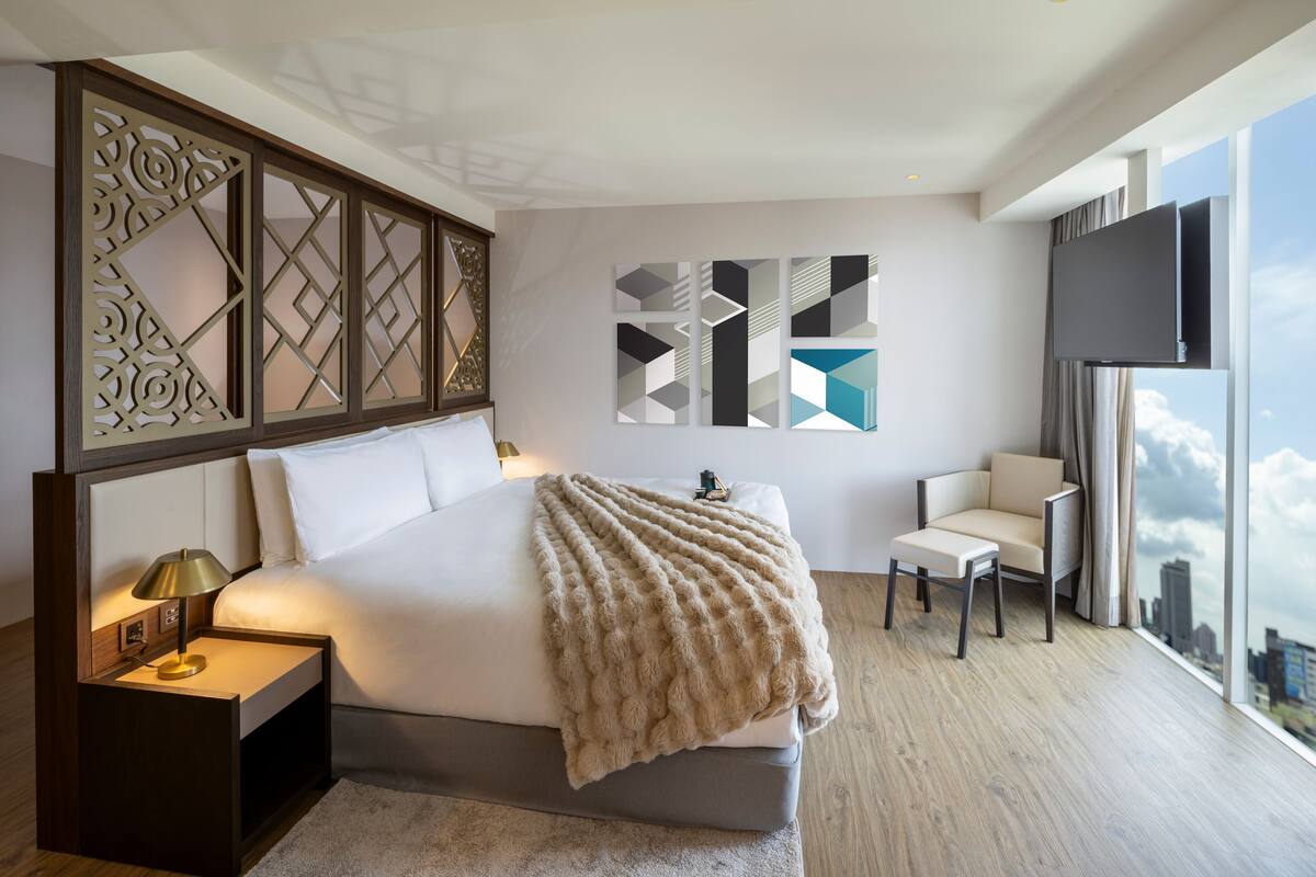 Quarto de hotel moderno com uma cama grande, uma mesa de cabeceira com abajur, uma cadeira e uma televisão.