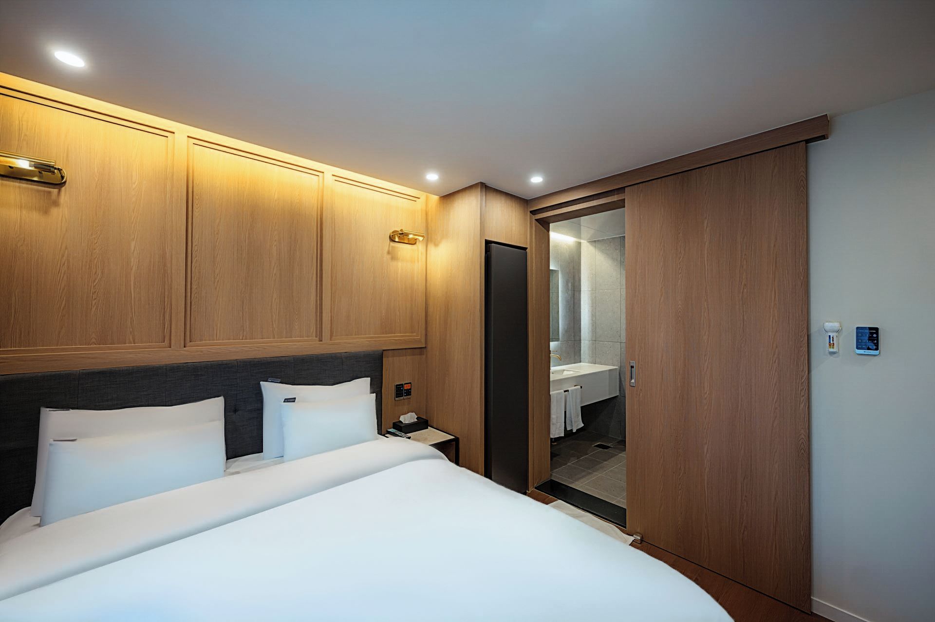 Quarto de hotel com uma cama grande, cabeceira de madeira, uma mesa de cabeceira e uma porta deslizante para um banheiro.