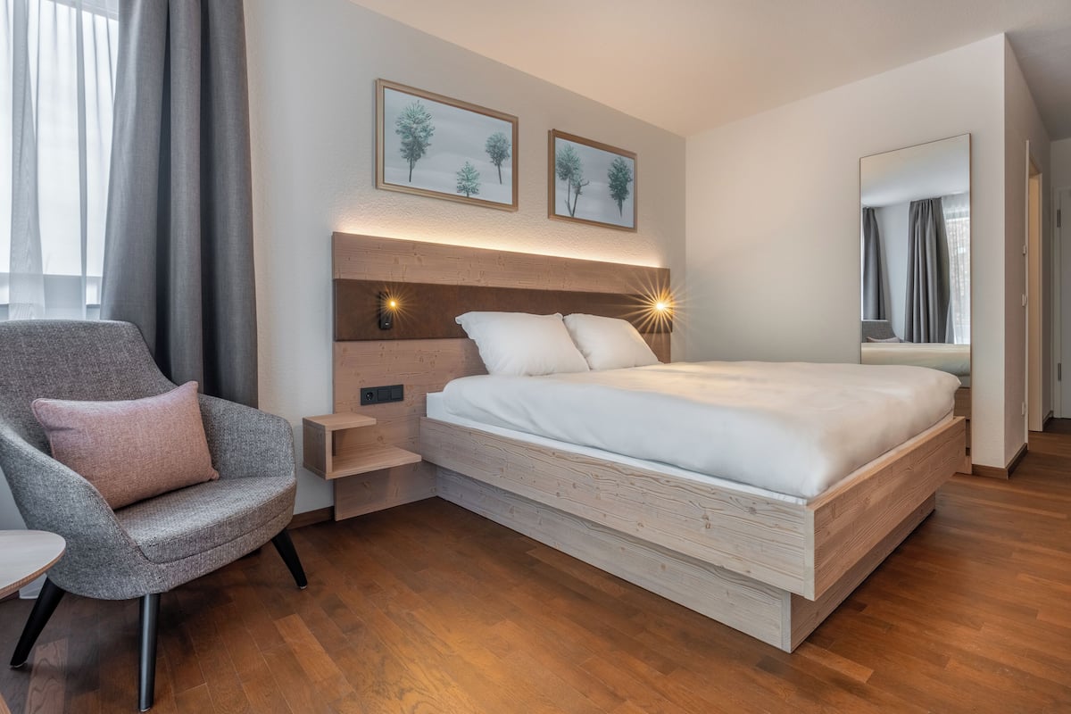 Quarto moderno com cabeceira de madeira, uma cama grande, uma poltrona cinzenta e duas molduras na parede.