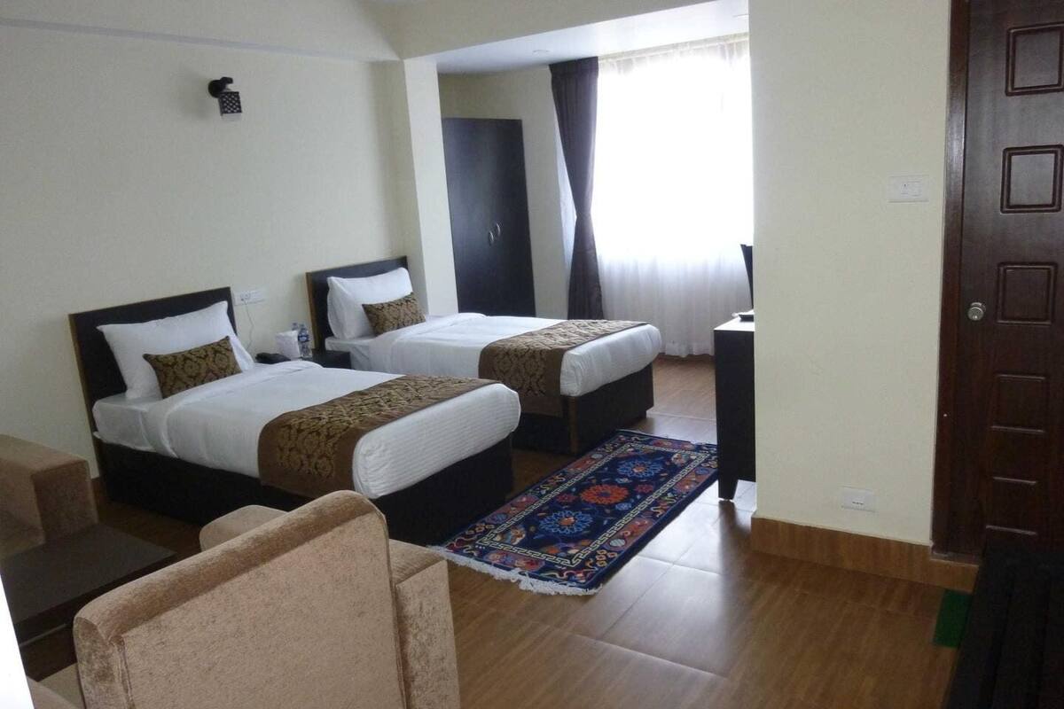Quarto de hotel com duas camas, uma pequena mesa, uma cadeira e uma janela com cortinas.