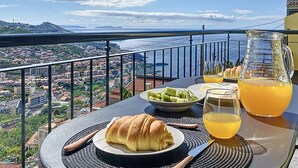 Uma mesa com croissants, uma travessa de frutas e um jarro de suco de laranja, com vista para o horizonte urbano e a água.