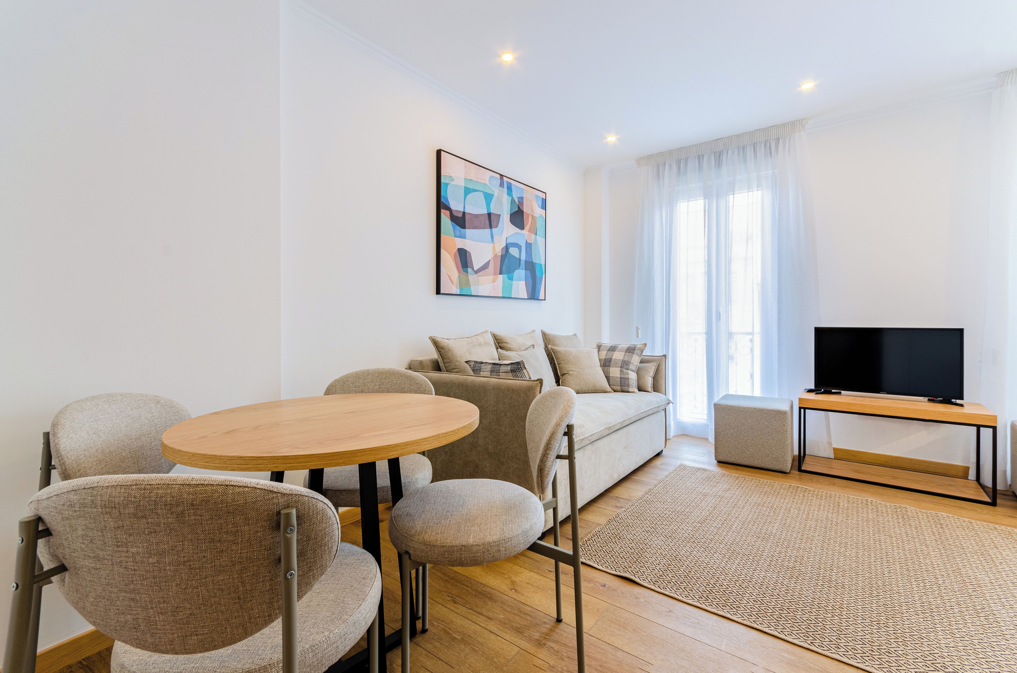 Uma sala de estar moderna com um sofá, duas poltronas, uma mesa redonda de madeira, uma TV em um móvel e um quadro na parede.
