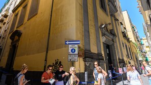Rua estreita com edifícios amarelos, placa indicando área para pedestres e uma barreira de corda azul.