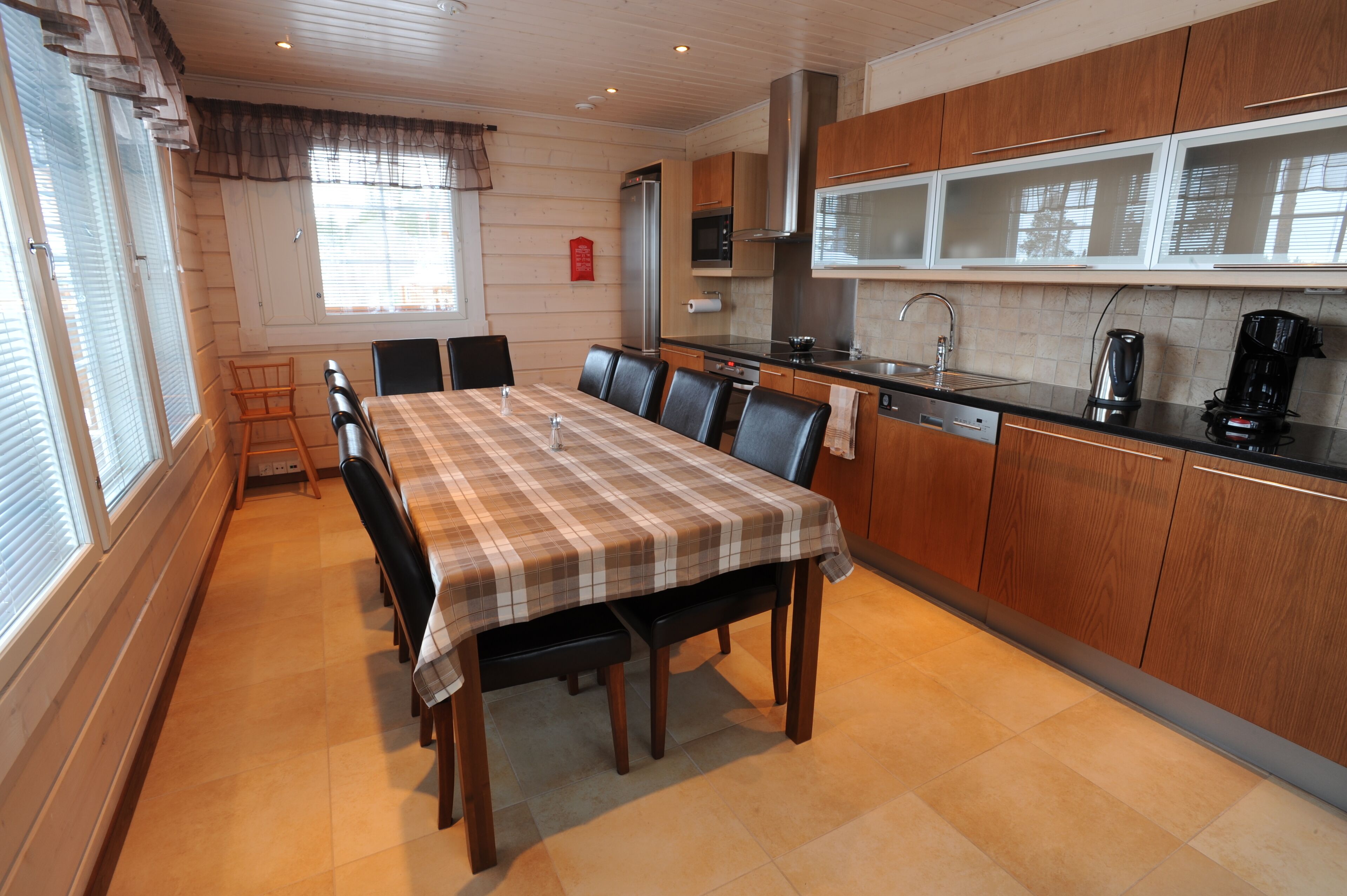 Uma sala de jantar com uma mesa posta para quatro pessoas, armários de madeira, uma cozinha com micro-ondas e uma janela com venezianas.