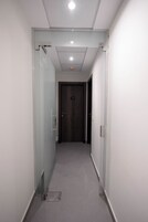 Um corredor estreito e bem iluminado, com um painel de vidro em um lado, uma porta no final e um respiradouro de metal no chão.