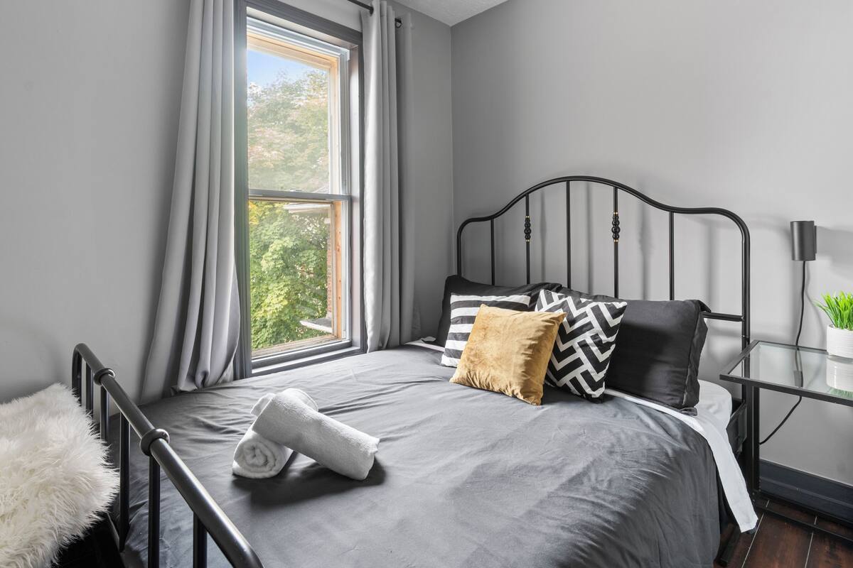 Uma cama bem arrumada com um espalha cama cinza, um travesseiro branco fofo e uma toalha branca enrolada. Também se vê uma cabeceira de metal preto e uma pequena mesa de cabeceira com uma planta.