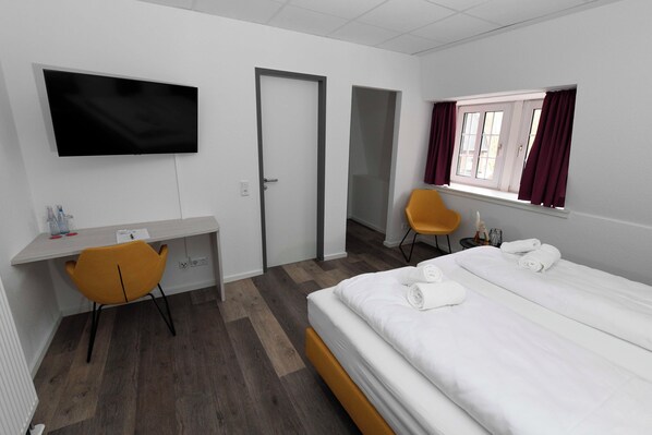 Quarto de hotel com cama, escrivaninha, cadeira, televisão e janela com cortinas.