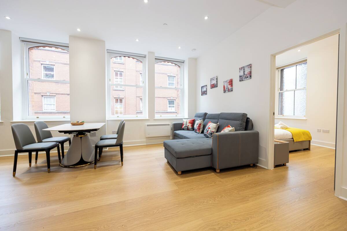 Sala de estar moderna com um sofá cinzento, uma mesa de jantar com cadeiras e grandes janelas com vista para um edifício de tijolos.