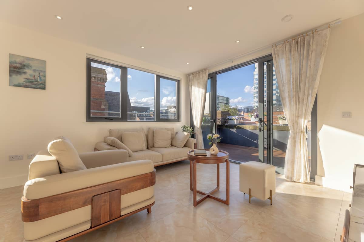 Uma sala de estar moderna com um sofá, uma mesa lateral de madeira redonda e vista para a cidade através de grandes janelas.