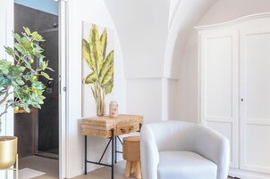 Um quarto com uma poltrona de cor clara, uma mesa lateral de madeira com um vaso, e uma planta grande.
