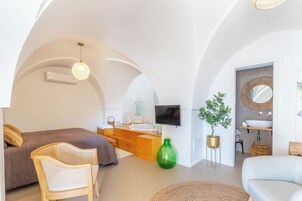 Quarto moderno com cama, poltrona, televisão, um vaso verde e banheiro com pia e espelho.