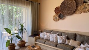 Uma sala de estar com sofá, almofadas estampadas, um banquinho trançado e uma peça de arte em tecido fixada na parede.