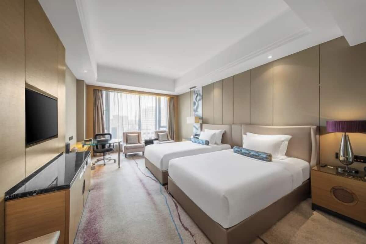 Quarto de hotel com duas camas, uma mesa, uma cadeira, uma TV e uma janela com cortinas.