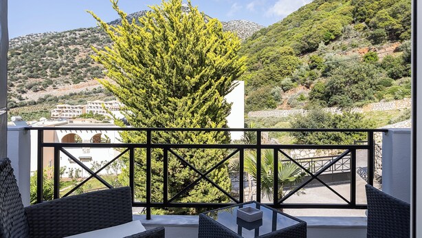 Uma varanda com móveis de vime, uma mesa de vidro e vista para uma paisagem montanhosa com vegetação e edifícios ao fundo.