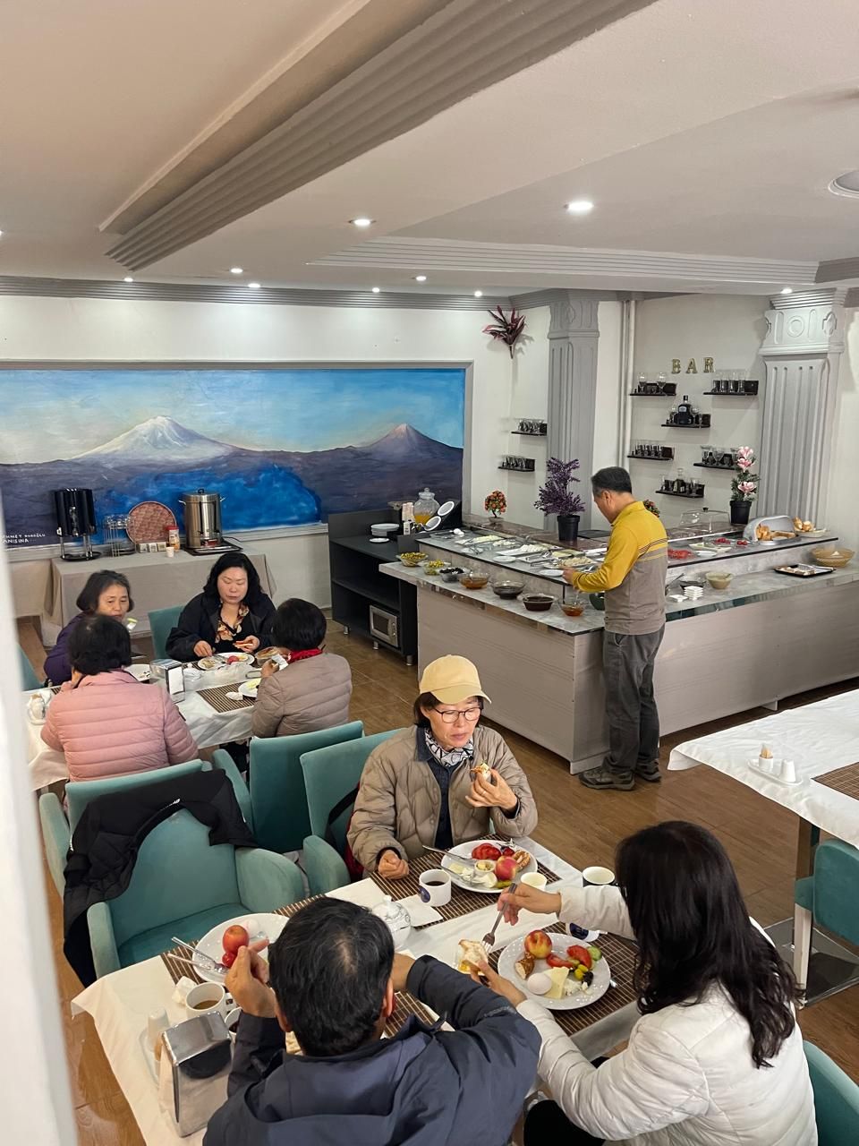 Restaurante com buffet, área de bar e pintura de uma montanha na parede.