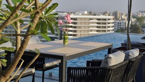Área à beira da piscina com mesa e cadeiras, um vaso com flores e vista para o horizonte urbano.