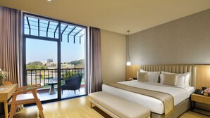 Quarto de hotel com uma cama grande, uma escrivaninha, um banco, uma cadeira e vista para o exterior por uma porta de correr.