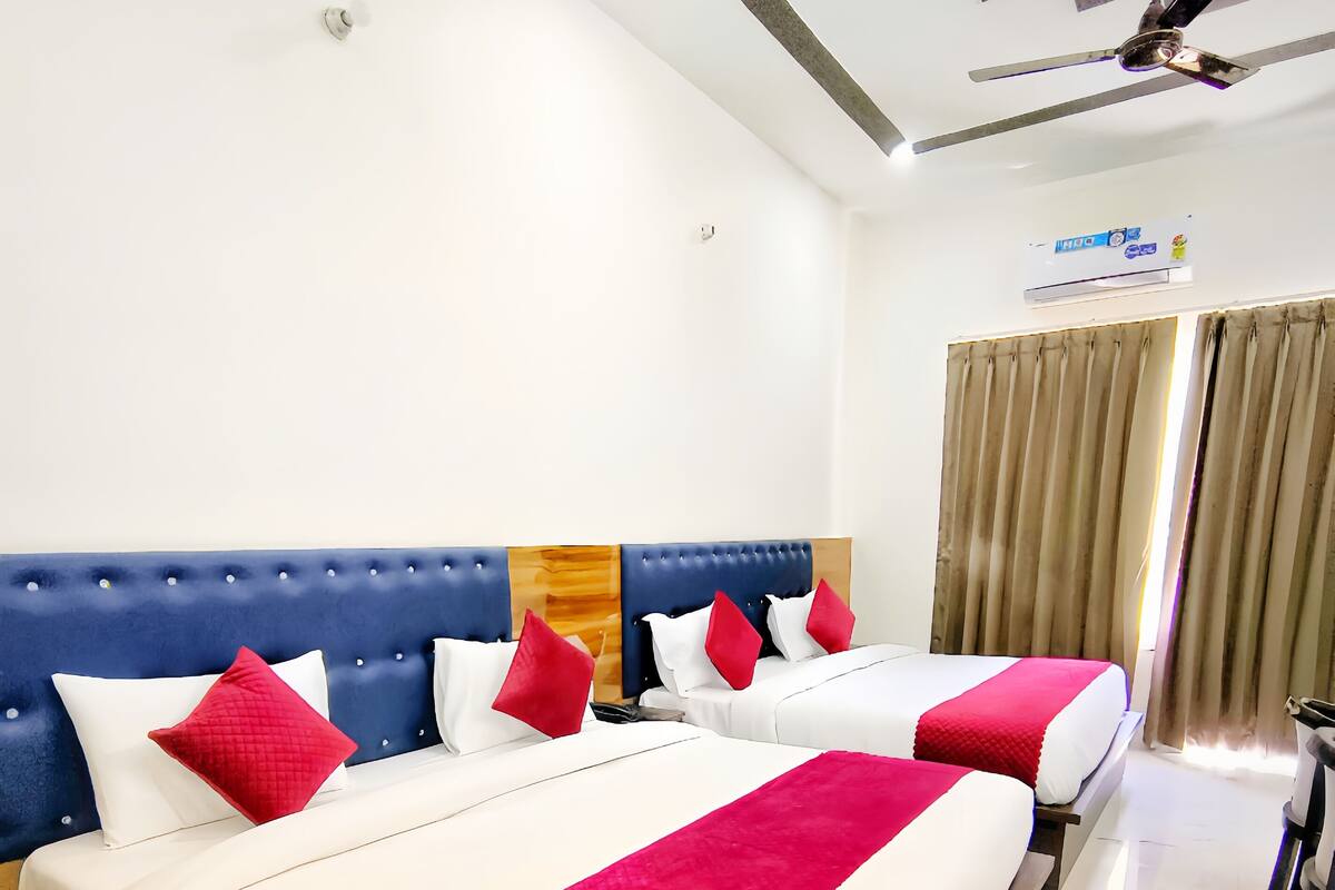 Quarto de hotel com duas camas, cabeceira azul, almofadas vermelhas e brancas, manta vermelha, ventilador de teto e janela com cortinas.